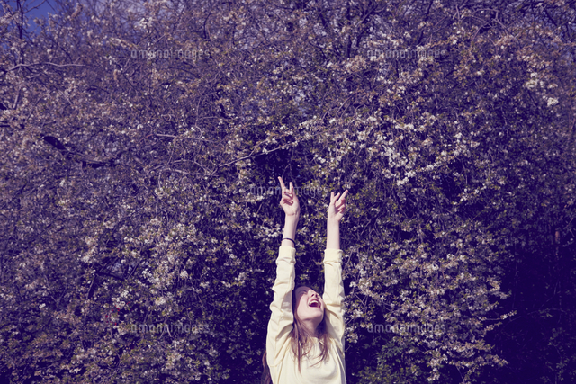 Portrait of girl in front of foliage with arms raised making peace sign ...