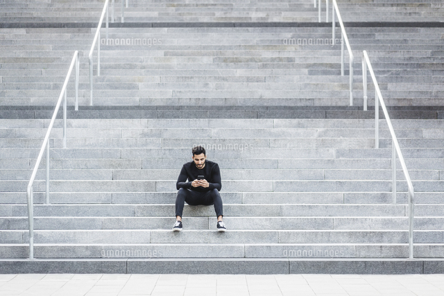 Young man using mobile phone while sitting on staircase[11081030410]の写真 ...
