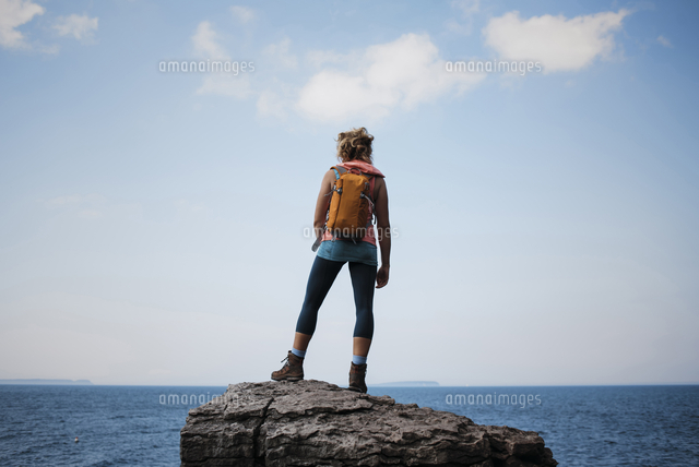 Rear view of female hiker with backpack standing on rock against sea ...
